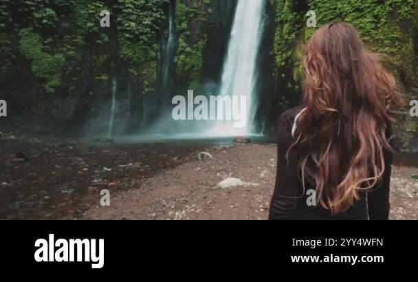A woman stands at the base of a majestic Bali waterfall, surrounded by ...