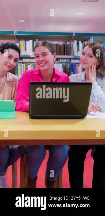 Three students in a library working together on a project with books and a laptop. They smile ...
