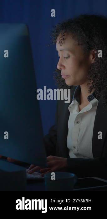 A determined woman in business attire focuses intently on her computer in a dim office ...