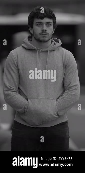 A young man stands in a thoughtful pose, hands placed in his pockets ...