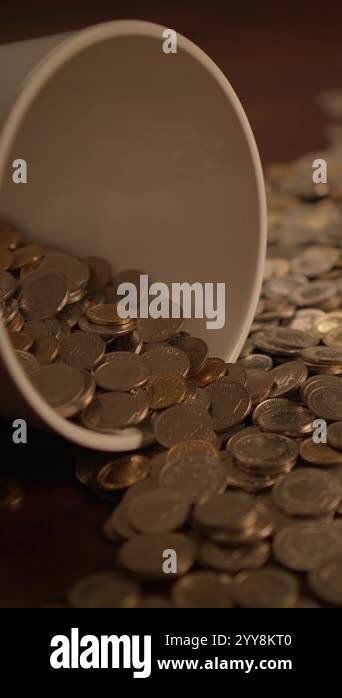 A white ceramic cup tipped over, spilling shiny coins onto a rustic ...