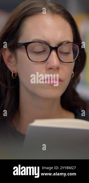 A focused young woman wearing glasses is intently engaged in reading a ...