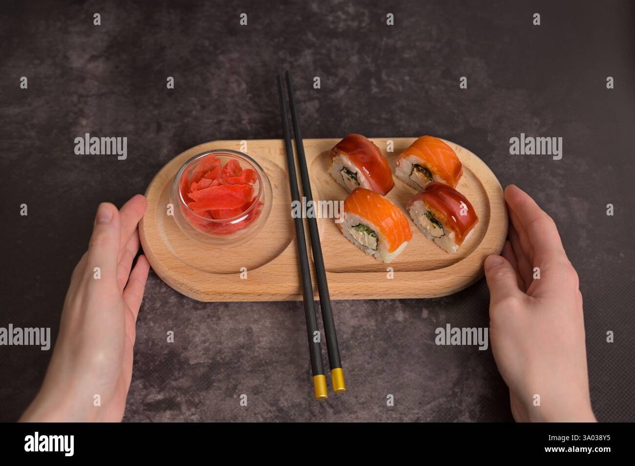 Woman's hands serve wooden serving board with set of sushi roll with rice and fish, chopsticks ...