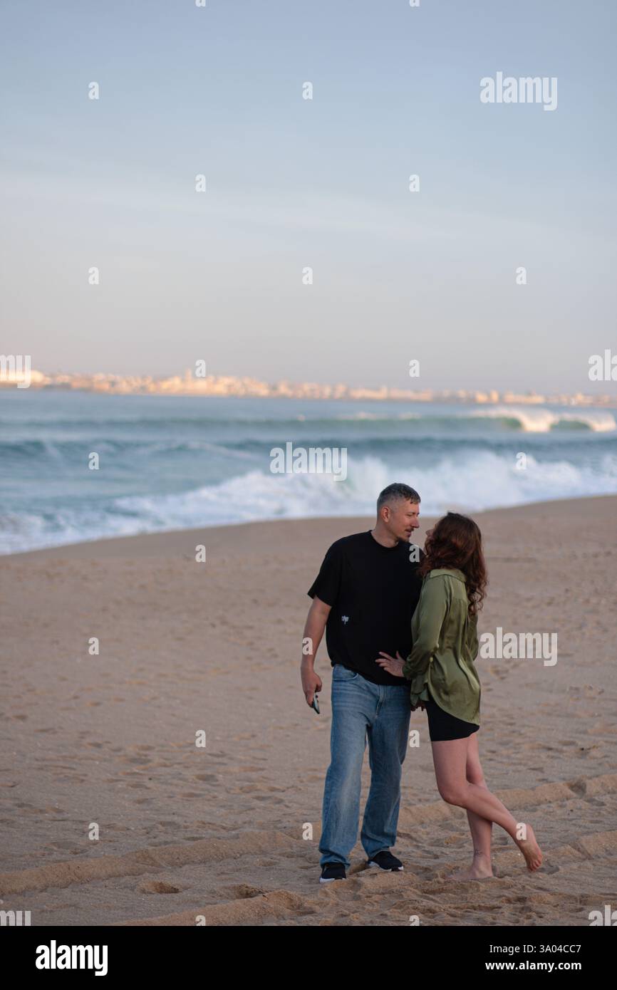 Young couple posing on ocean beach with waves and city on horizon ...