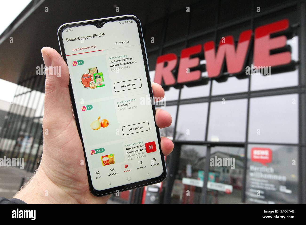 Ein Mann hält vor einem Rewe-Markt sein Smartphone in der Hand. Darauf ...