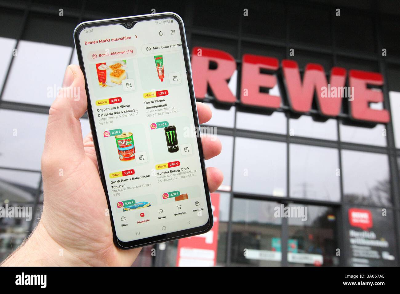 Ein Mann hält vor einem Rewe-Markt sein Smartphone in der Hand. Darauf ...