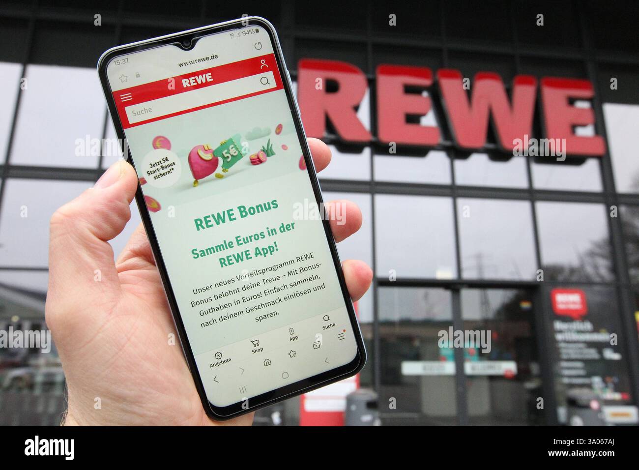 Ein Mann hält vor einem Rewe-Markt sein Smartphone in der Hand. Darauf ...
