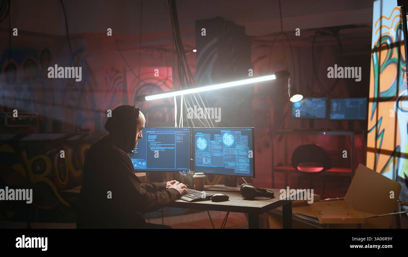 Hacker running phishing software on computer in criminals den with graffiti sprayed on walls ...