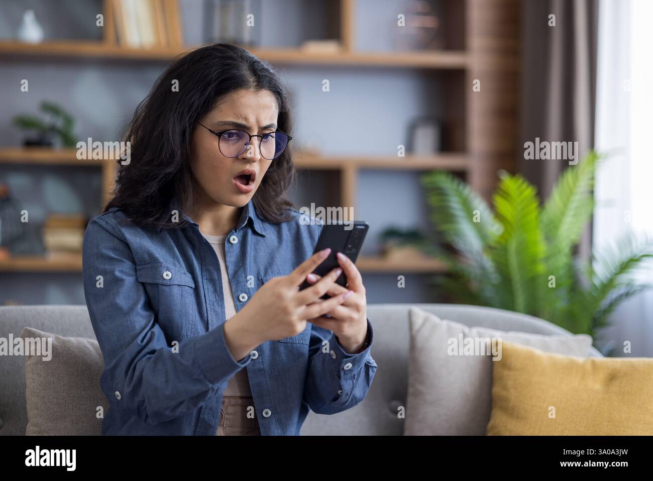 A young woman sits on her couch and stares in shock at her smartphone ...
