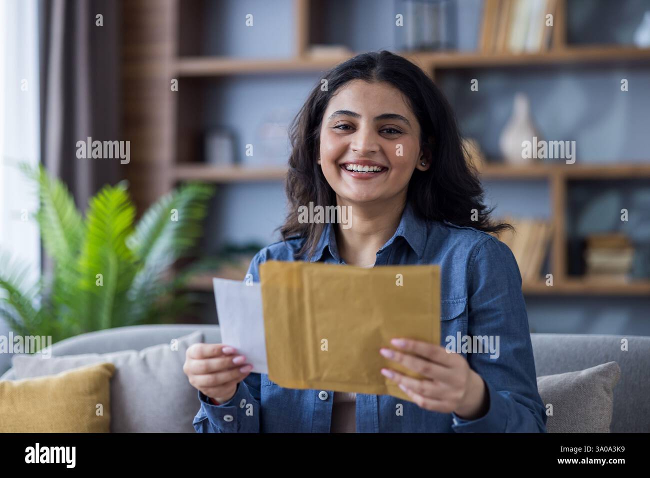 A young woman with a beaming smile sits on her couch, holding an ...