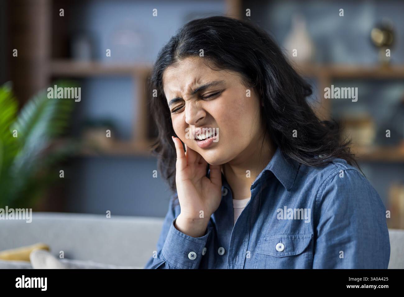 A young woman sits at home, wearing a blue shirt and suffering from a ...