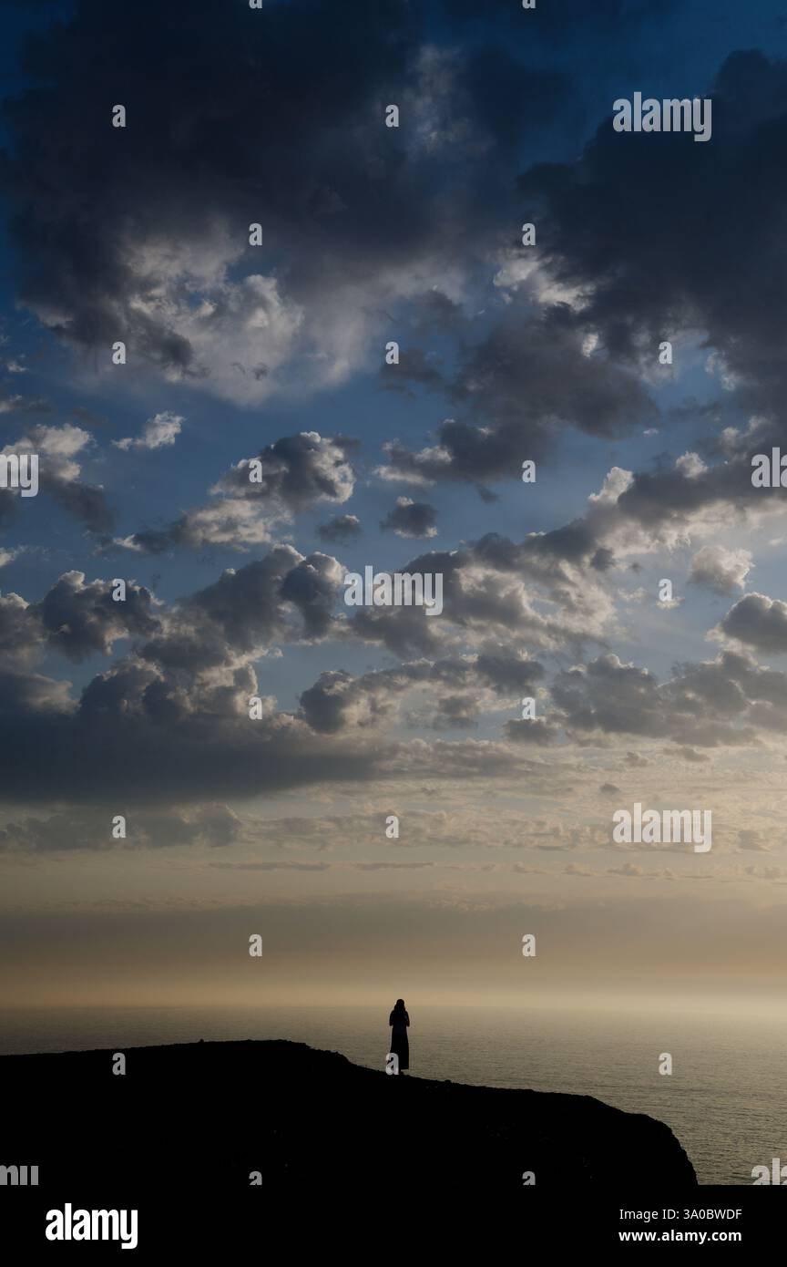 Lone figure stands on a coastal cliff under a dramatic sky at sunset in ...