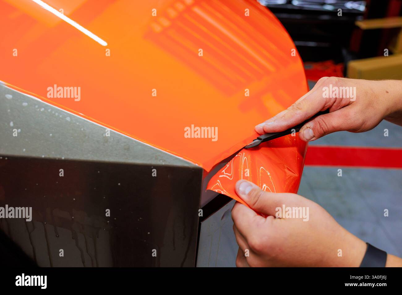 Car worker skillfully applies an orange vinyl wrap to car rear panel ...