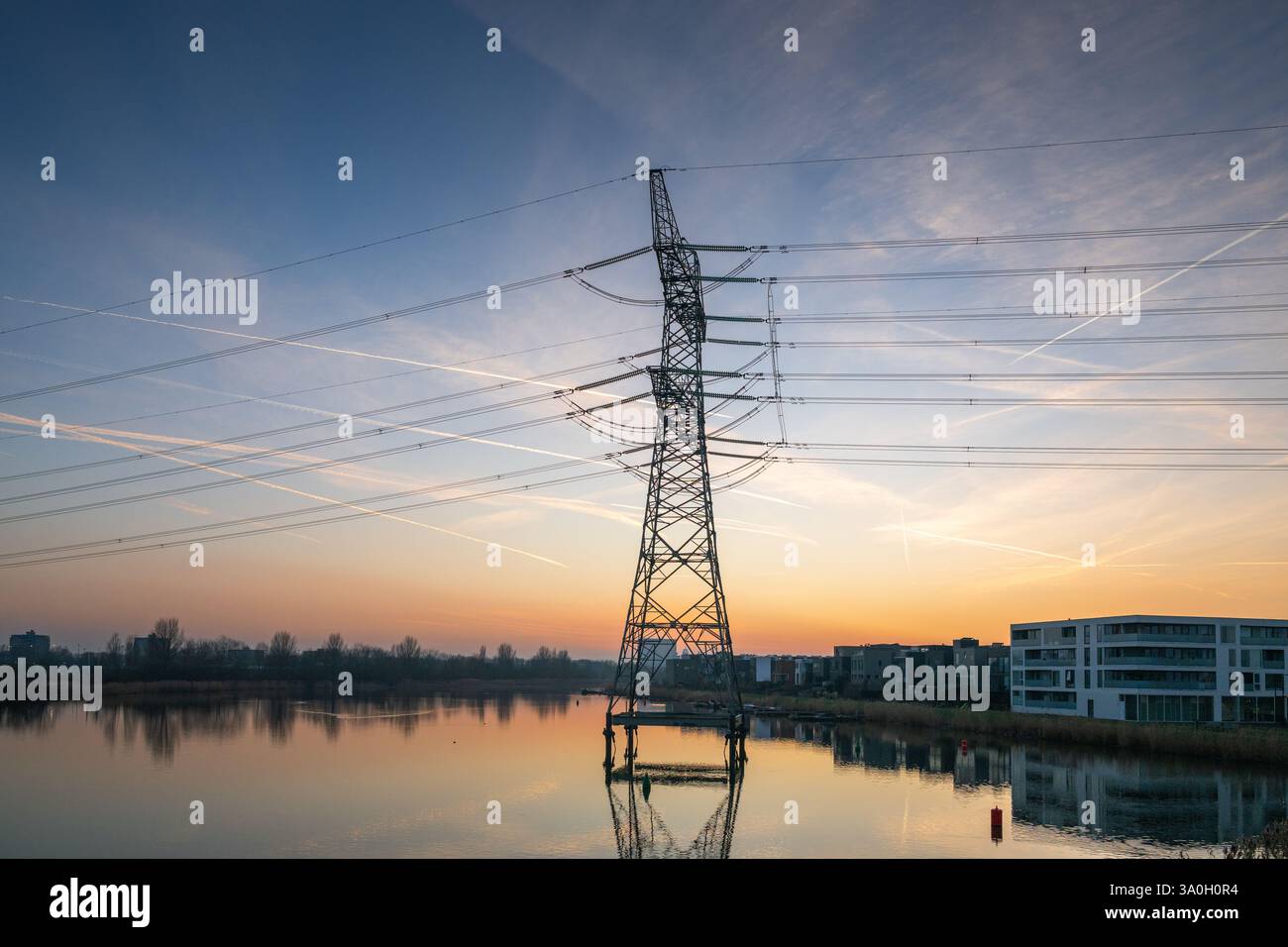 High voltage power line tower over water at sunset in a modern urban ...