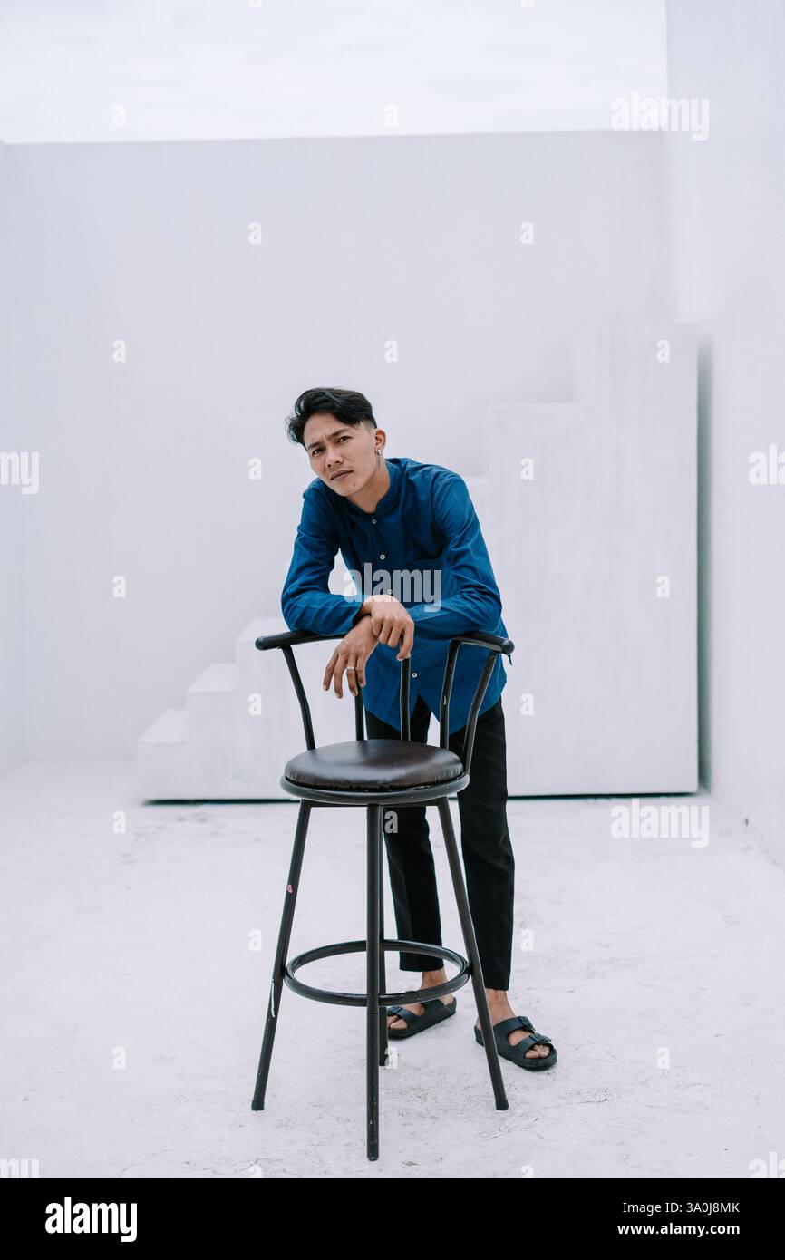 A portrait of a young asian man casually leaning on a stool in a bright ...