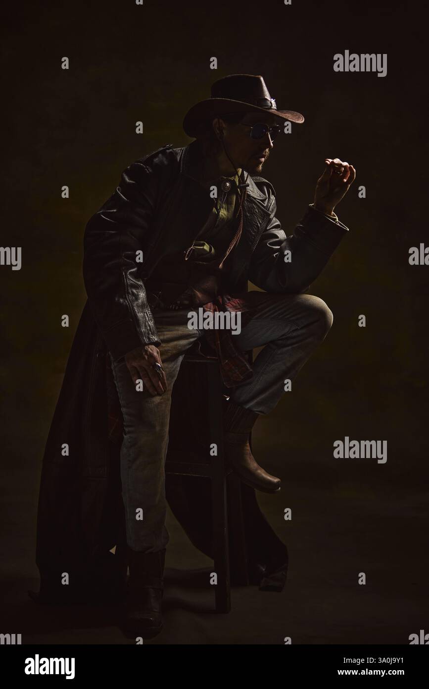 Brutal man, cowboy sitting on stool in dark leather coat, holding ...