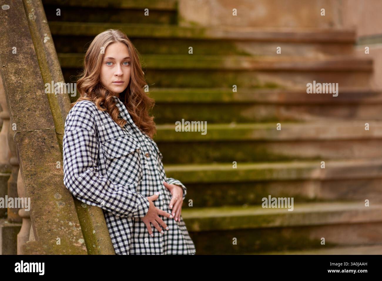 A young woman with wavy light brown hair and blue eyes leans against an ...