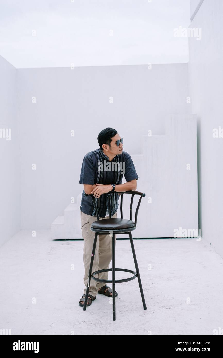 A man in casual stylish attire leans pensively on a chair within a ...