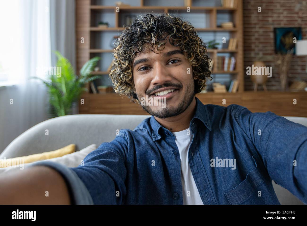 A smiling man with curly hair takes a selfie at home, casually dressed ...