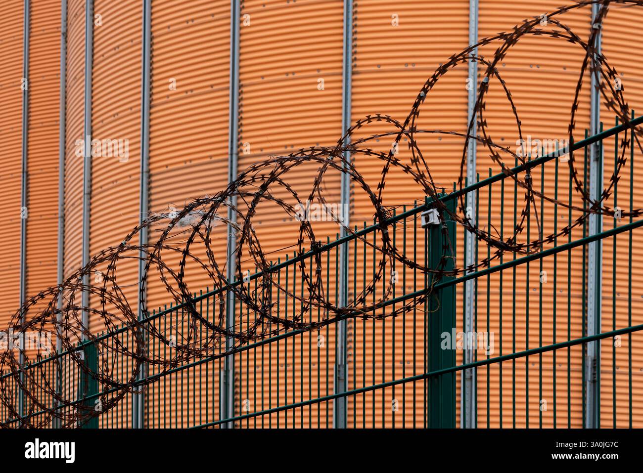 A barbed wire fence stands prominently in front of a large industrial ...