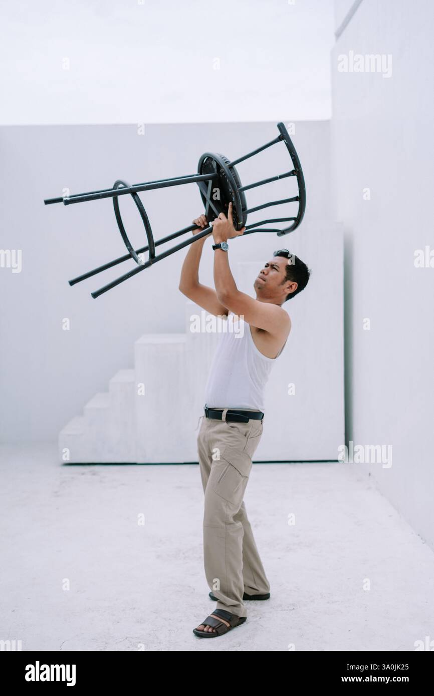 A man struggles to lift a black chair overhead in a bright, minimalist ...