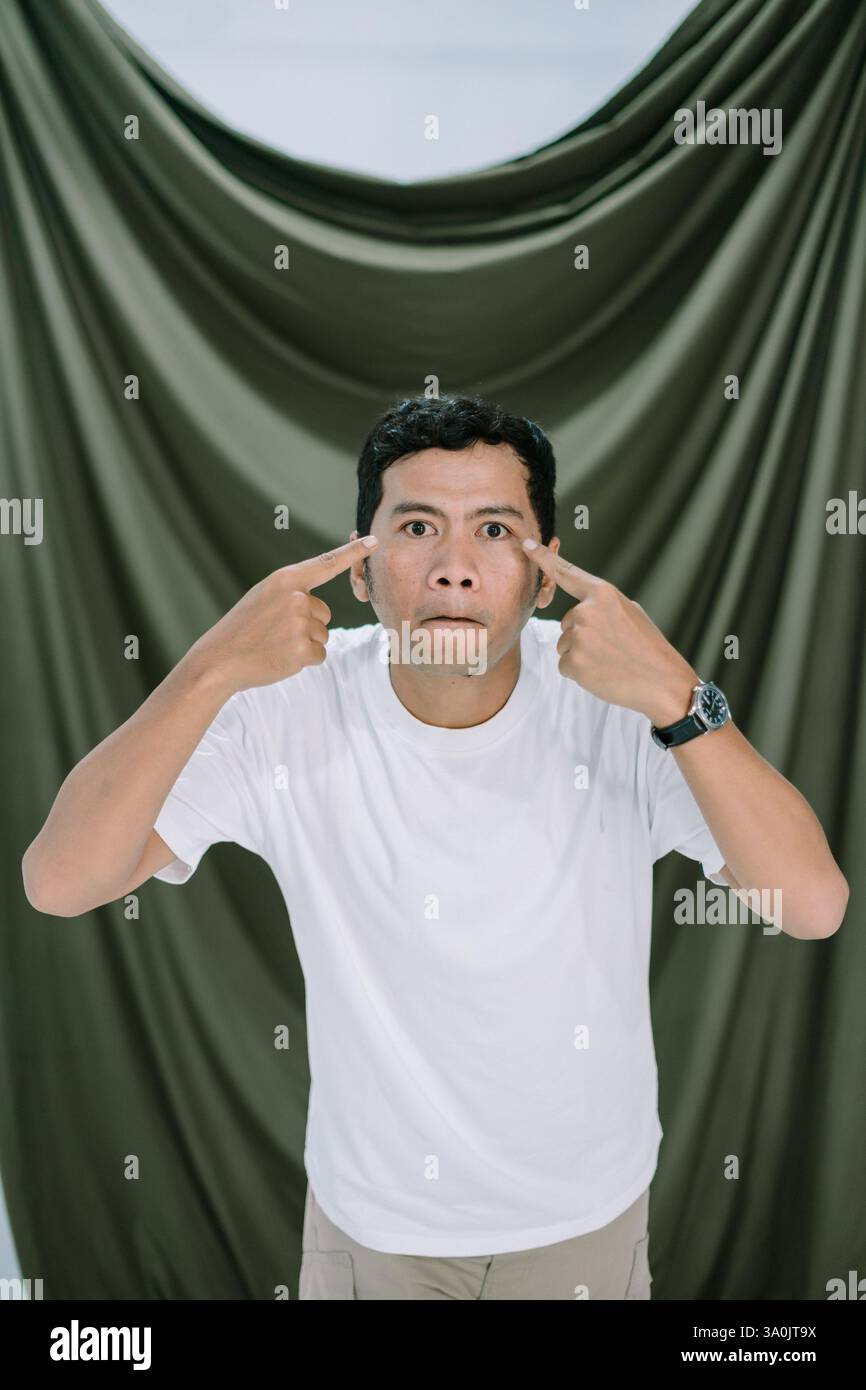 An Asian man points to his eyes with a confused expression, set against ...
