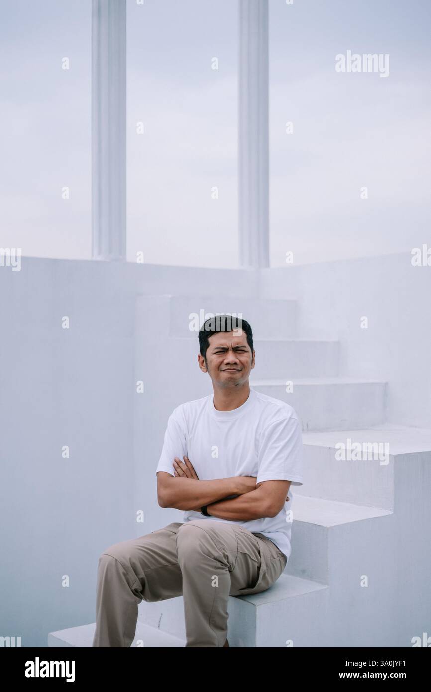 An asian man sits with crossed arms on a bright white, minimalist ...