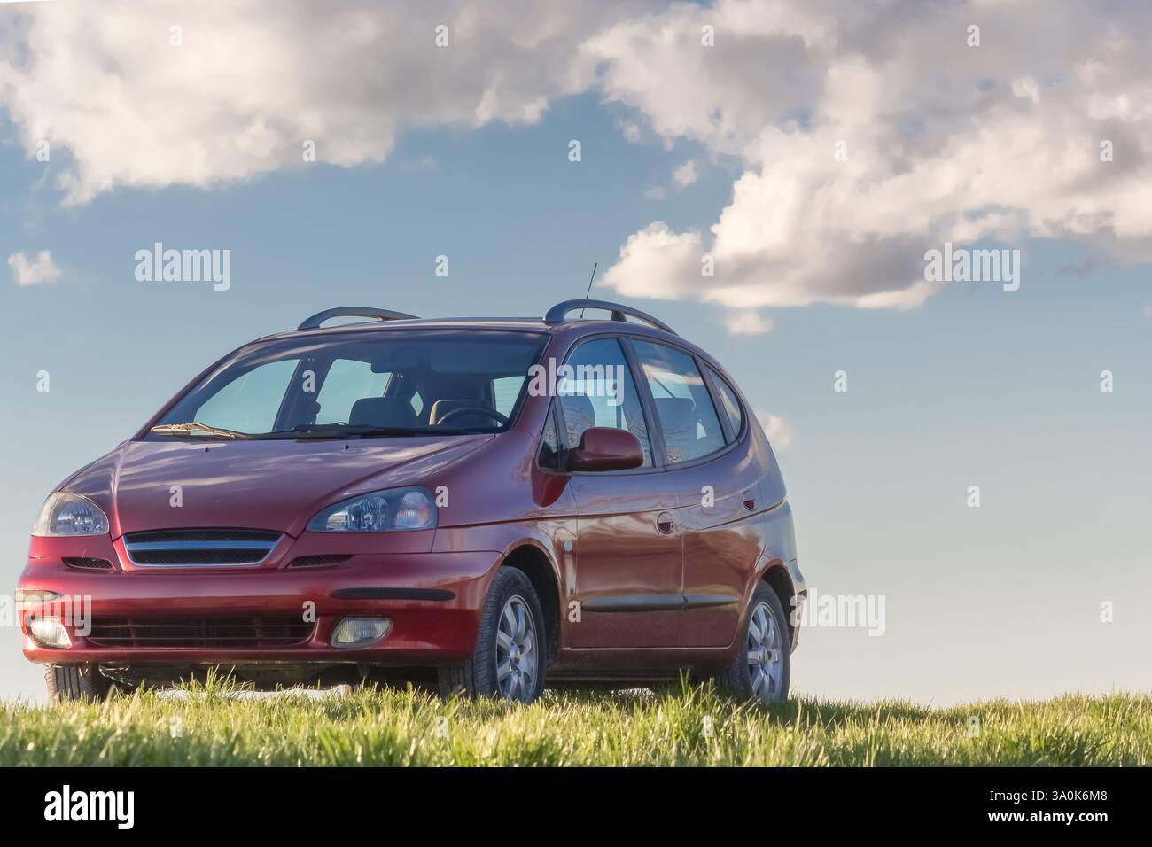 A compact red car is parked peacefully on a grassy field under a bright ...