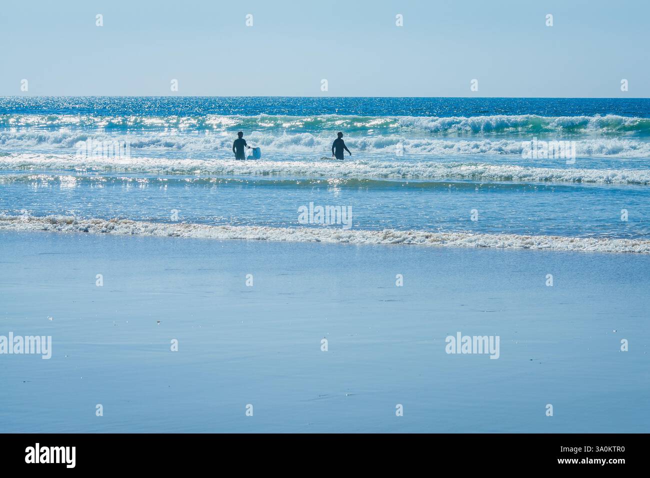 This image shows two surfers wading into the ocean, walking through the ...