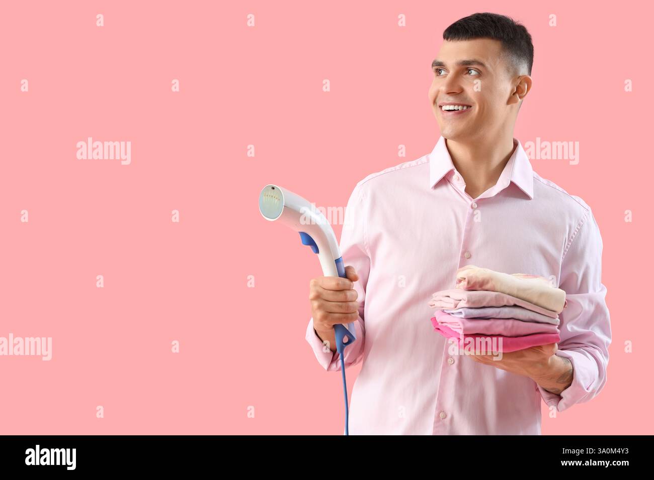 Happy young man with steamer and stack of clean clothes on pink ...