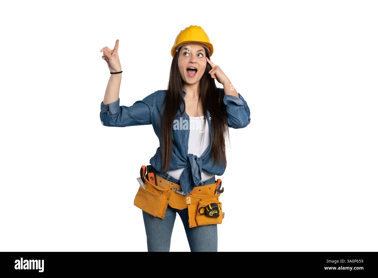 Smiling young woman in a yellow helmet and denim shirt with a tool belt, playfully pointing ...