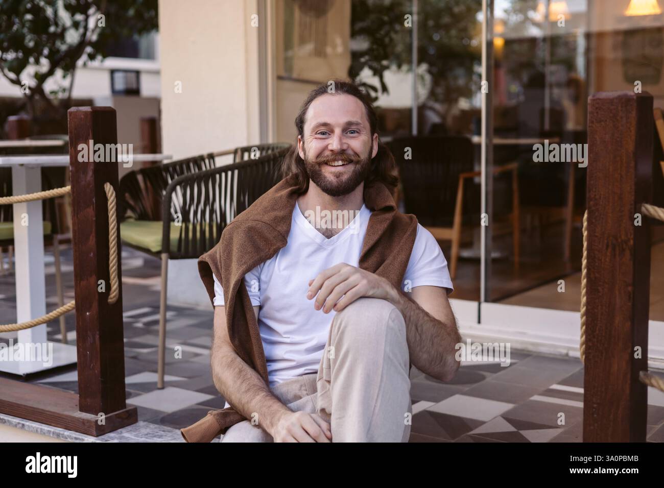 A man with long hair is relaxing on steps of cafe, smiling at the ...