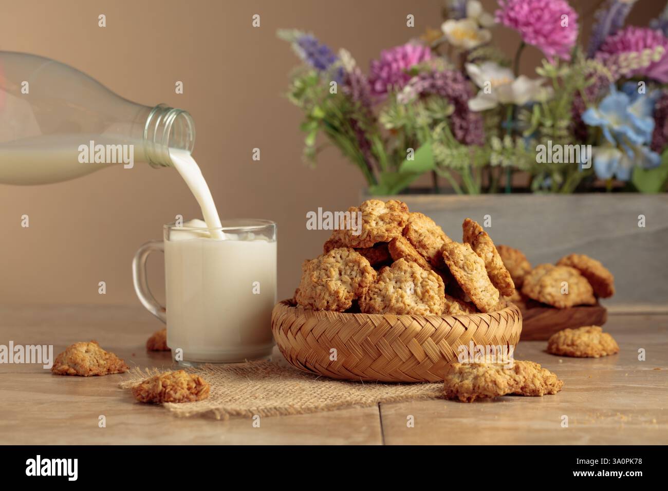 Oatmeal cookies and milk rest on a kitchen table. Milk is poured from a ...