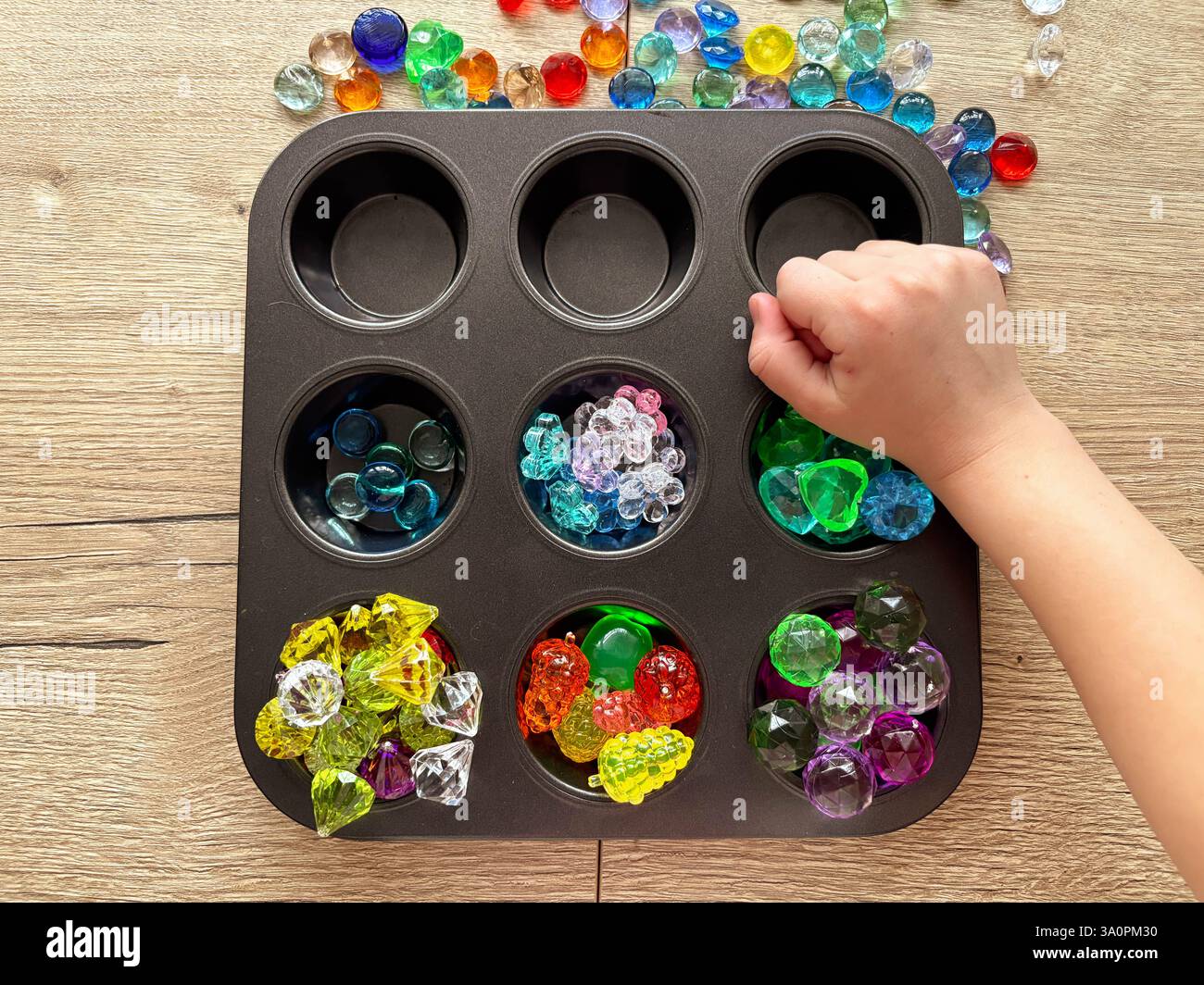 A child's hand arranges glass pebbles in a black metal mold. The child ...