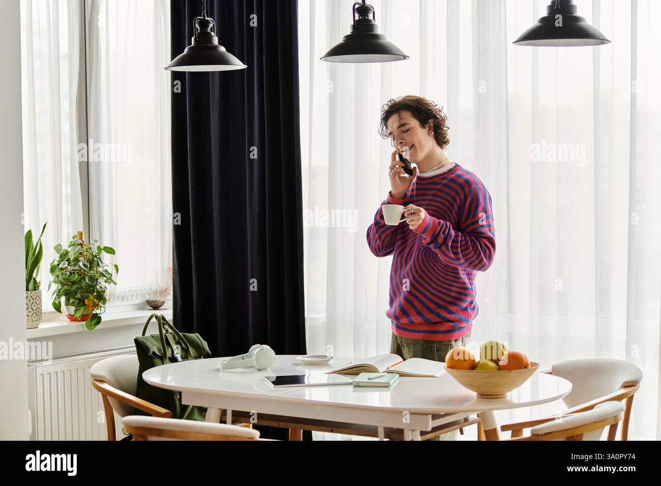 A young non-binary person talks on the phone while sipping tea in their ...