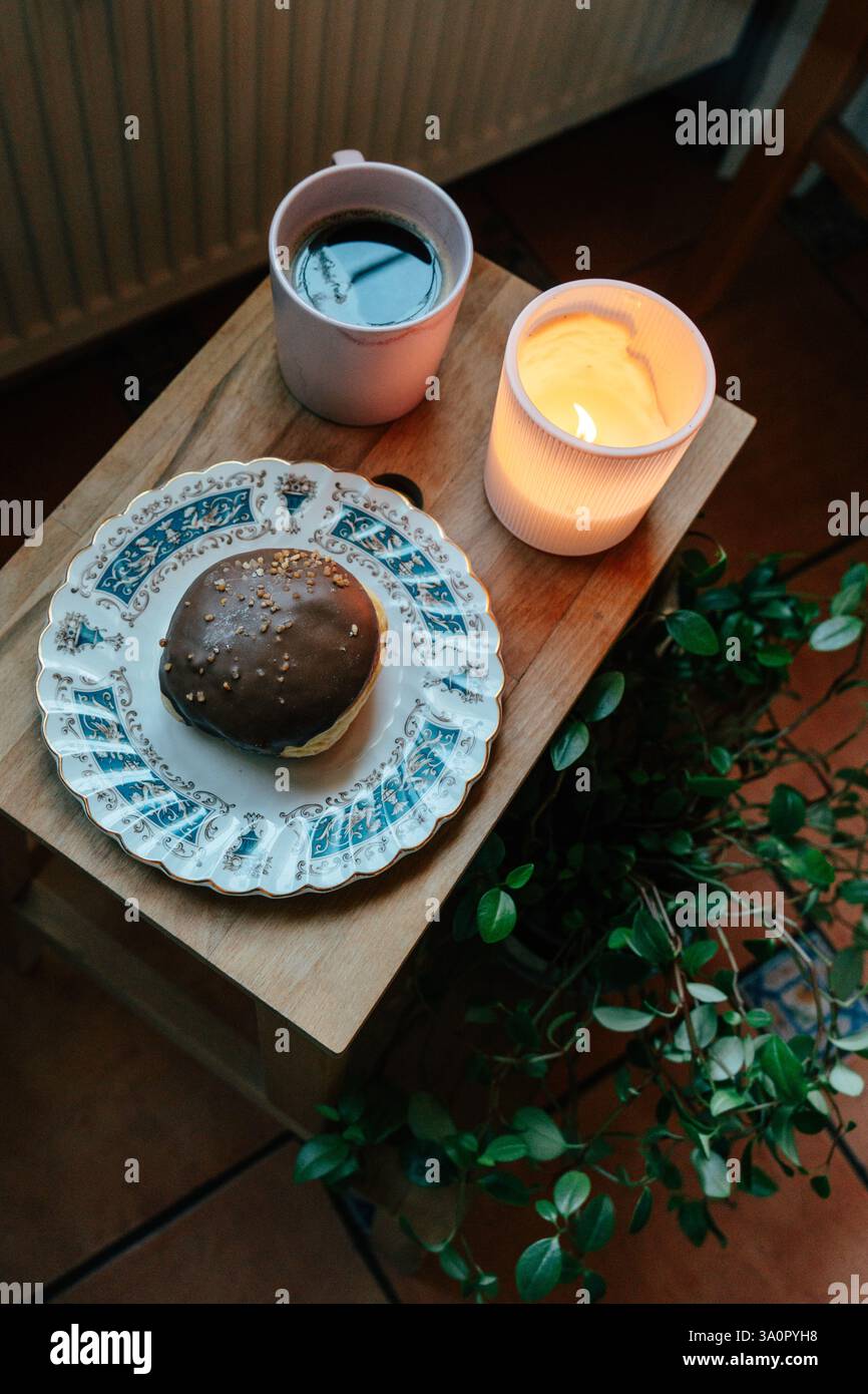 A dark pastry sits on an ornate plate next to a cup of coffee and a lit ...