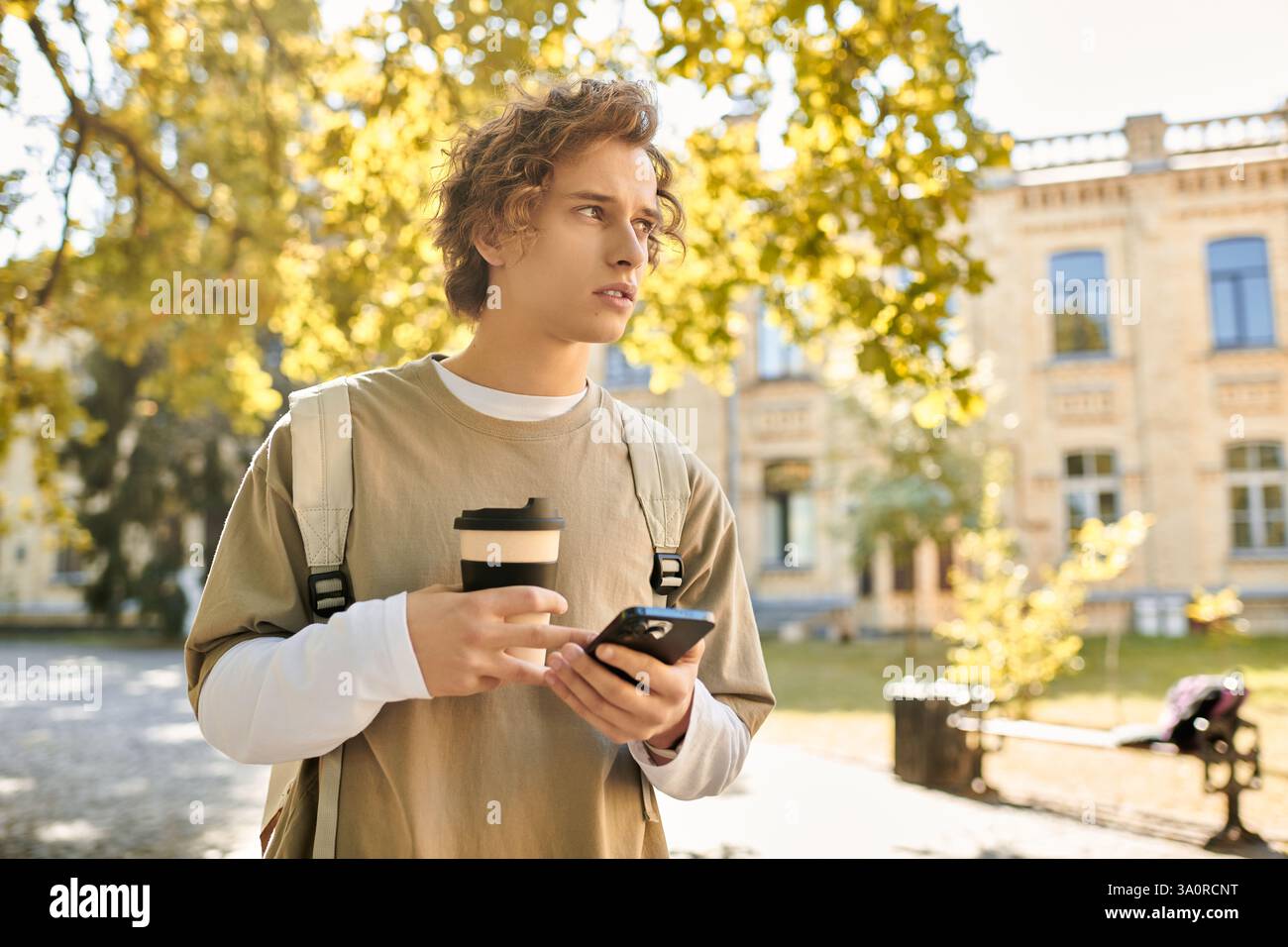 Young man stands outside in a sunny park, sipping coffee and engaged ...