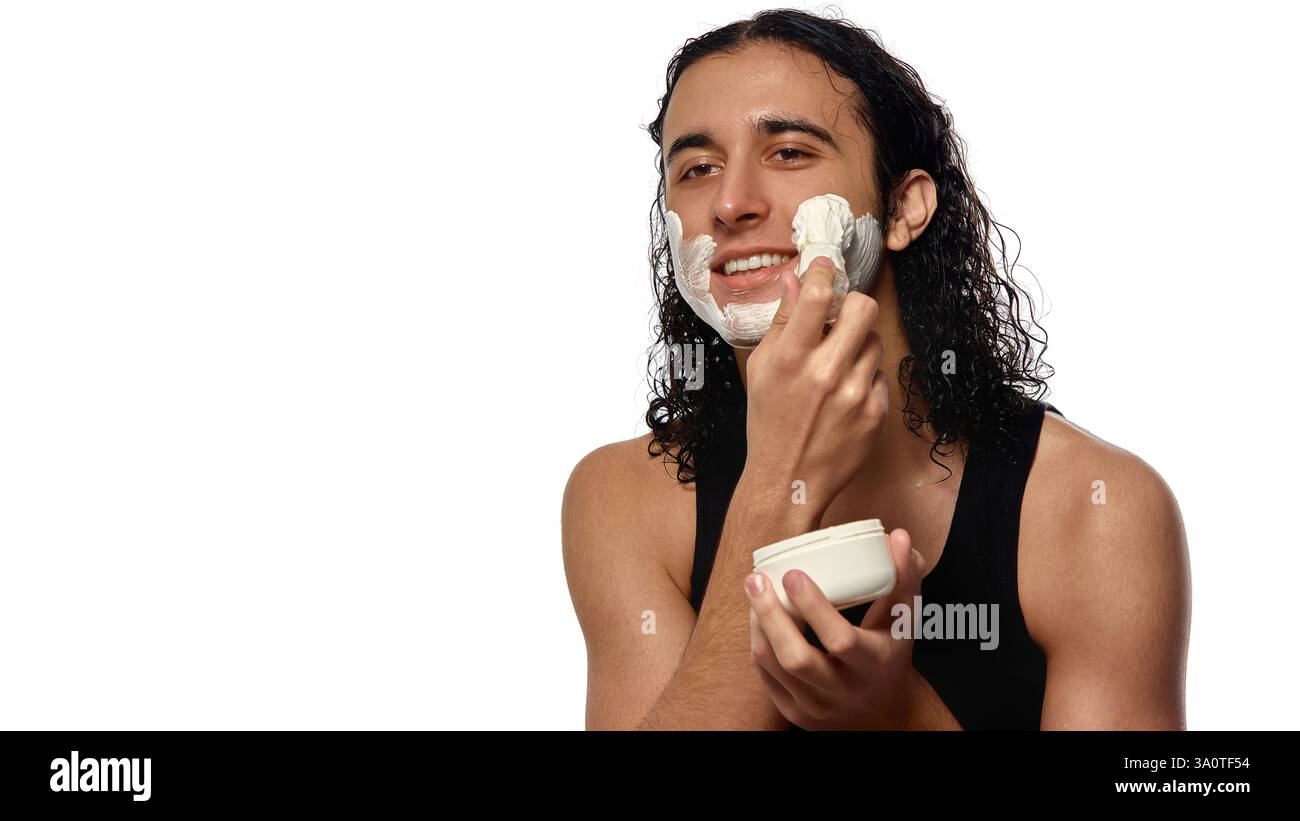 Smiling young man applying shaving cream to jawline with brush while holding cream jar against ...