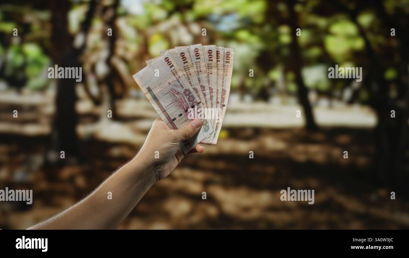 Man holding saudi riyal banknotes in a park setting, with trees forming ...