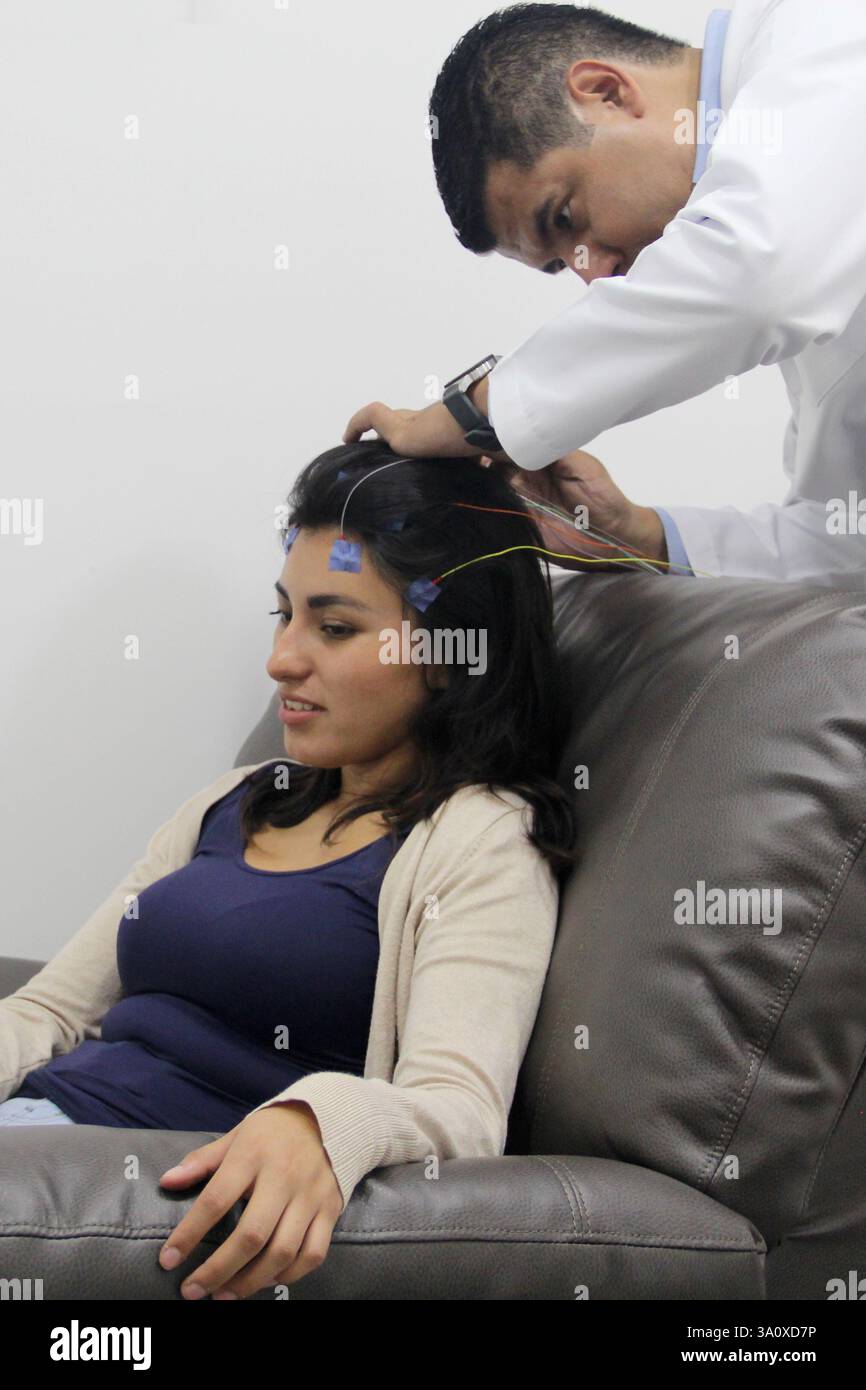 Dark-skinned Latino neurologist specialist doctor places electrodes on the head of a 20-year-old ...