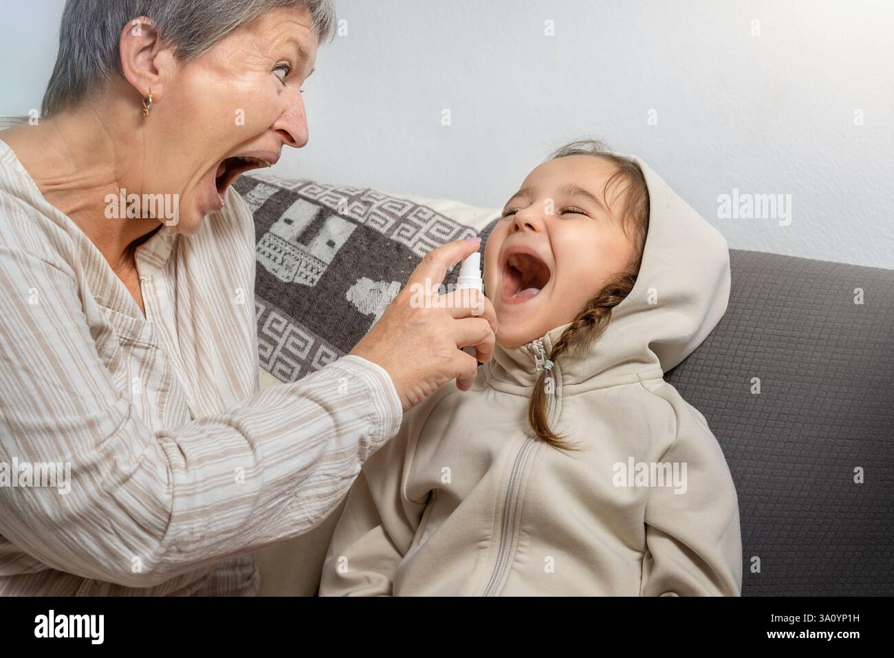 Senior woman spraying medicine into child's mouth while sitting on sofa ...