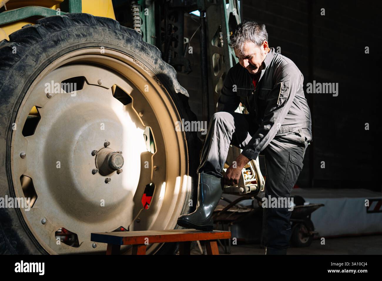 Caucasian male farmer putting on rubber boots next to a tractor wheel, getting ready for work in ...