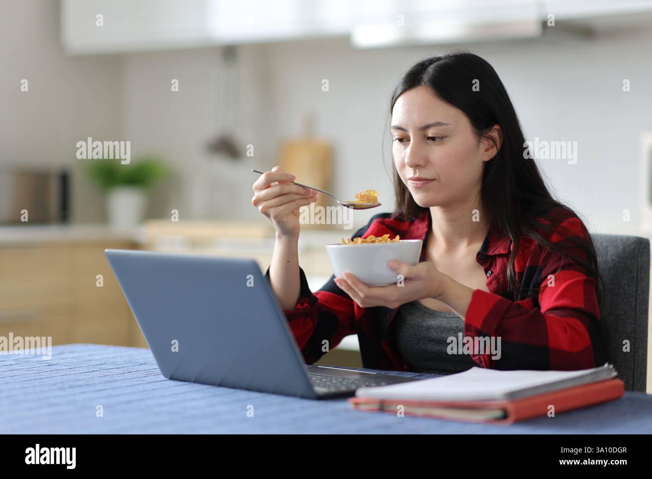 Asian student eating cereals using laptop sitting in the kitchan in a ...
