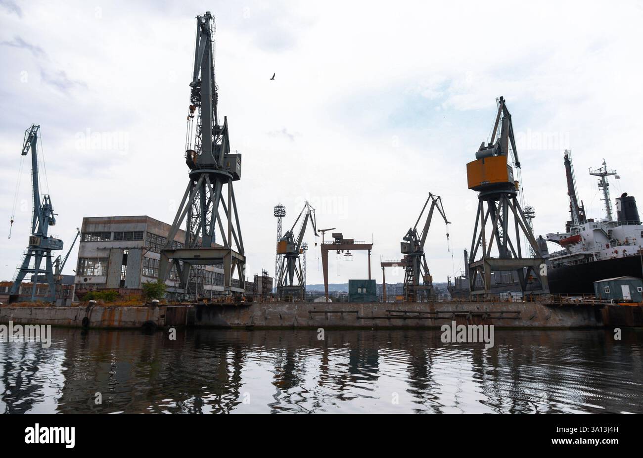 A busy industrial shipyard with large cranes, dockside buildings, and a ...
