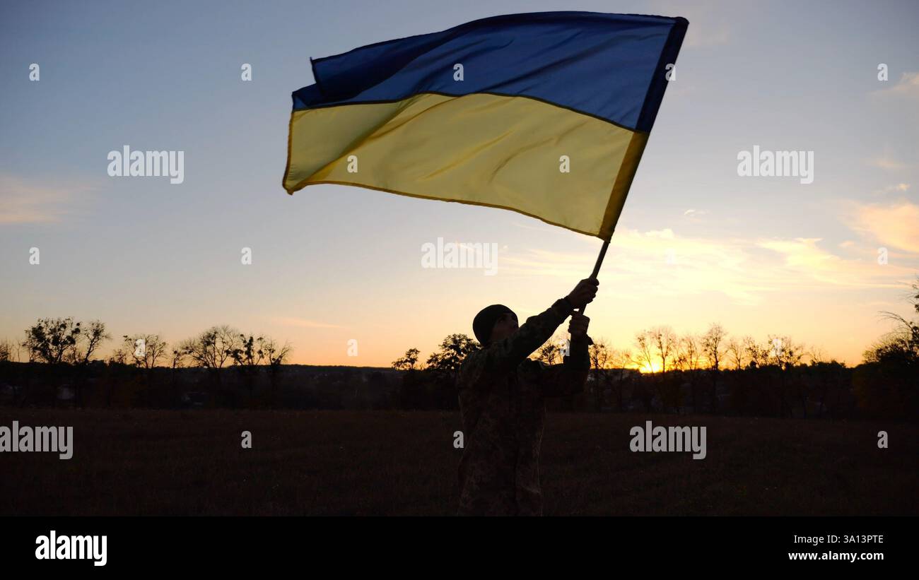 Male ukrainian army soldier going and waving flag of Ukraine on meadow ...