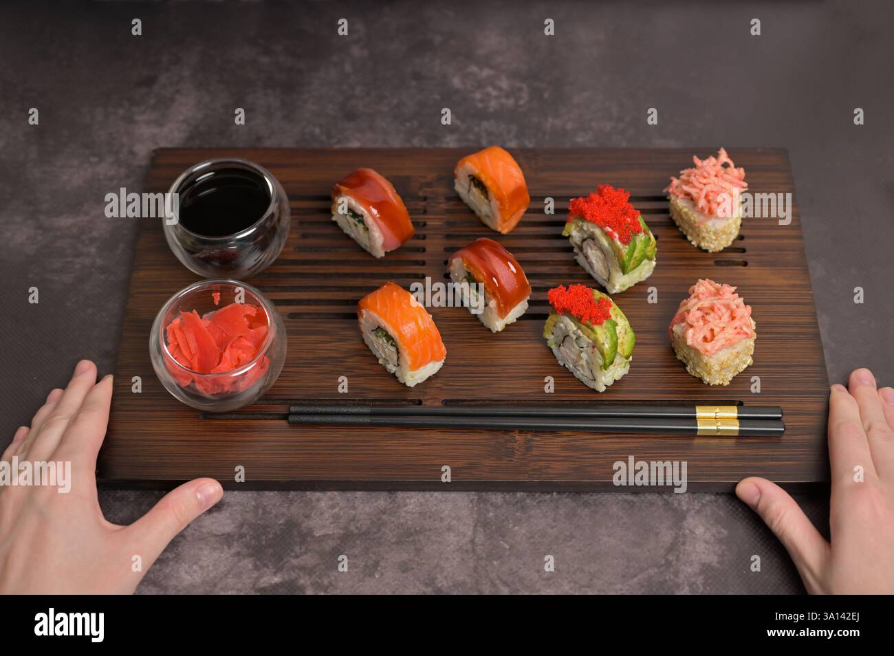 Woman's hands serve wooden serving board with set of sushi roll, ginger, soy sauce, chopsticks ...