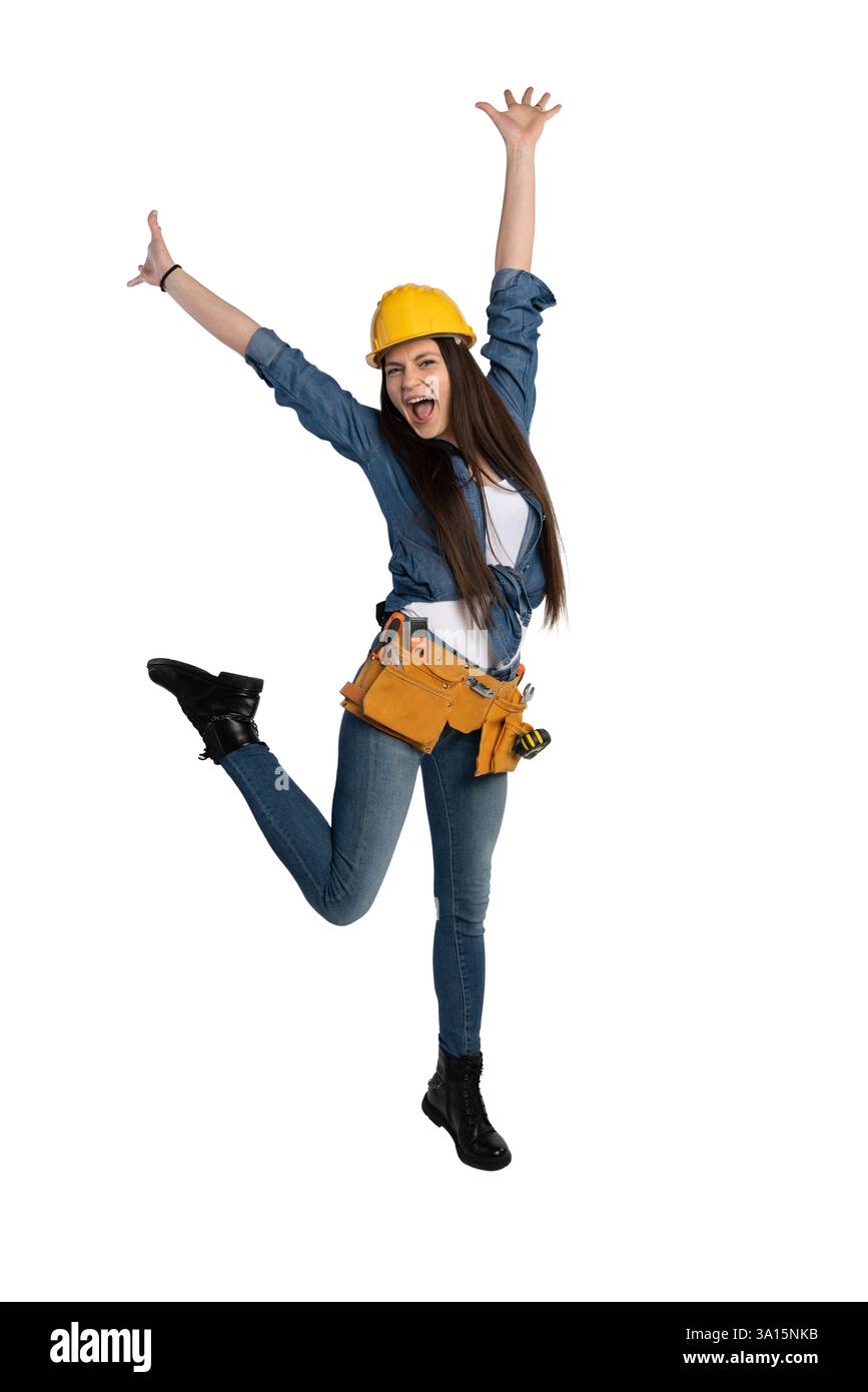 Young female construction worker wearing a yellow hard hat and tool belt, energetically jumping ...