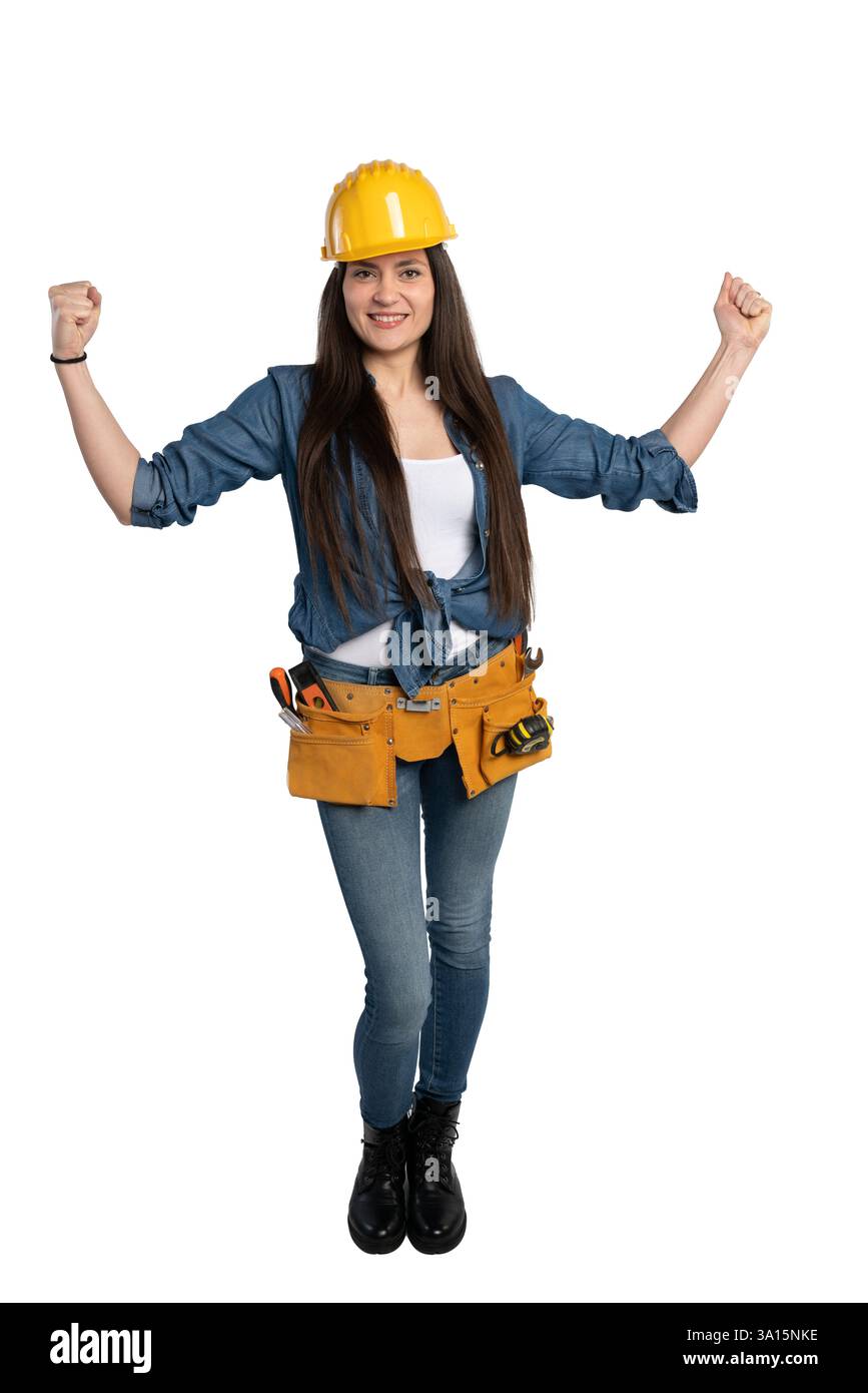 Young female construction worker wearing a yellow hard hat and tool belt, smiling while raising ...