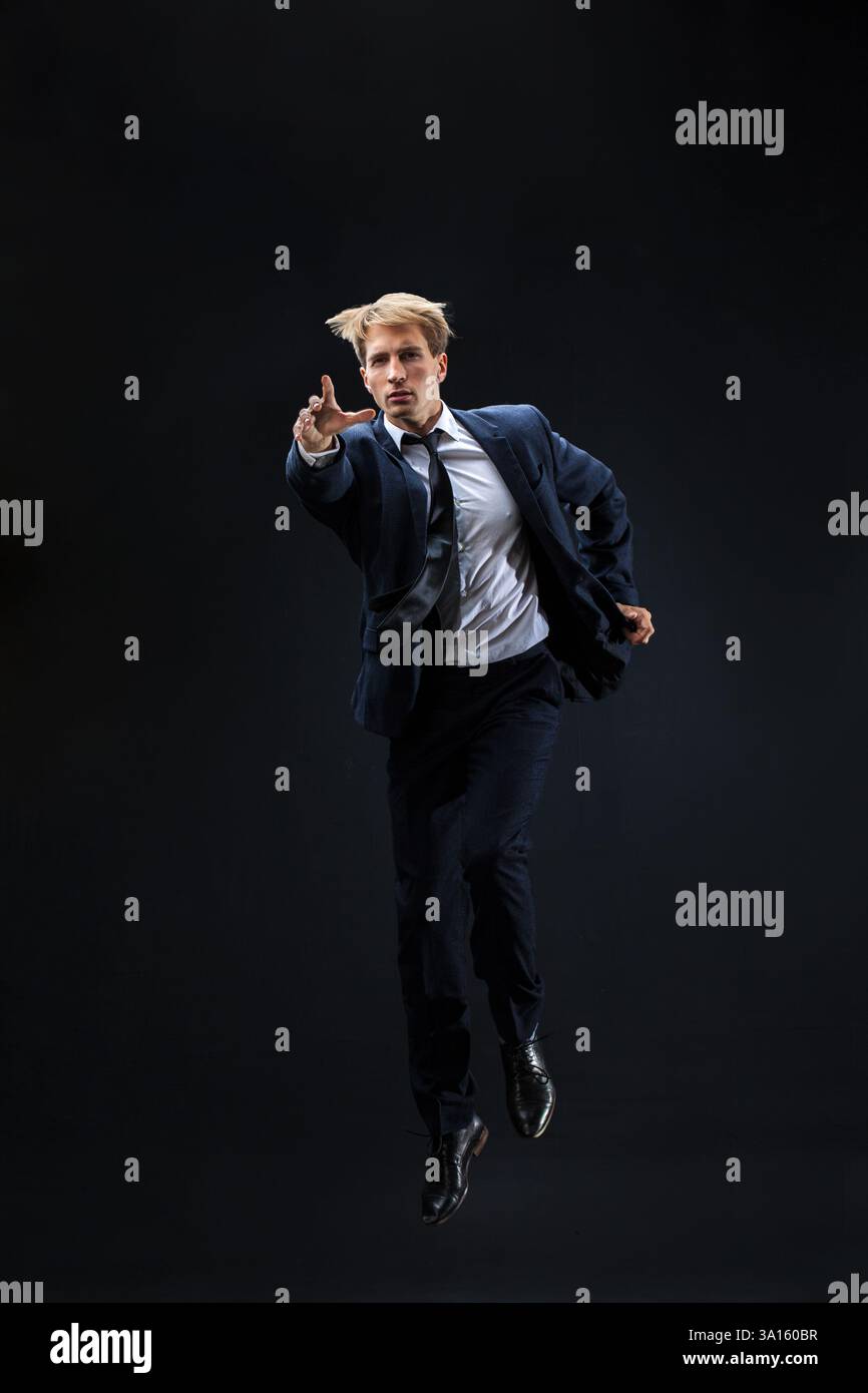 In a dark studio, a man in a sharp navy suit leaps mid-air, reaching ...