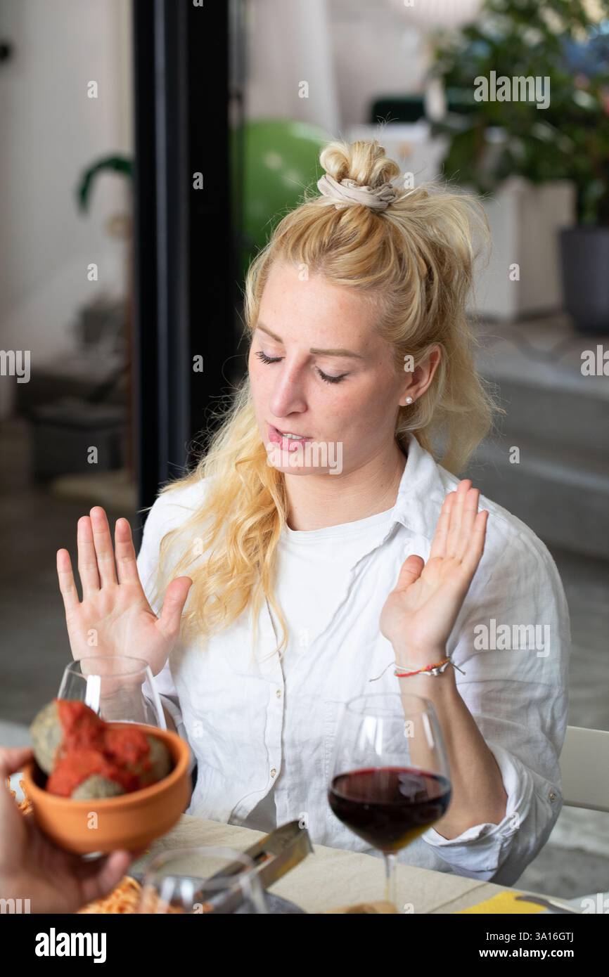 Blond woman raising her hands and refusing food at a restaurant table ...